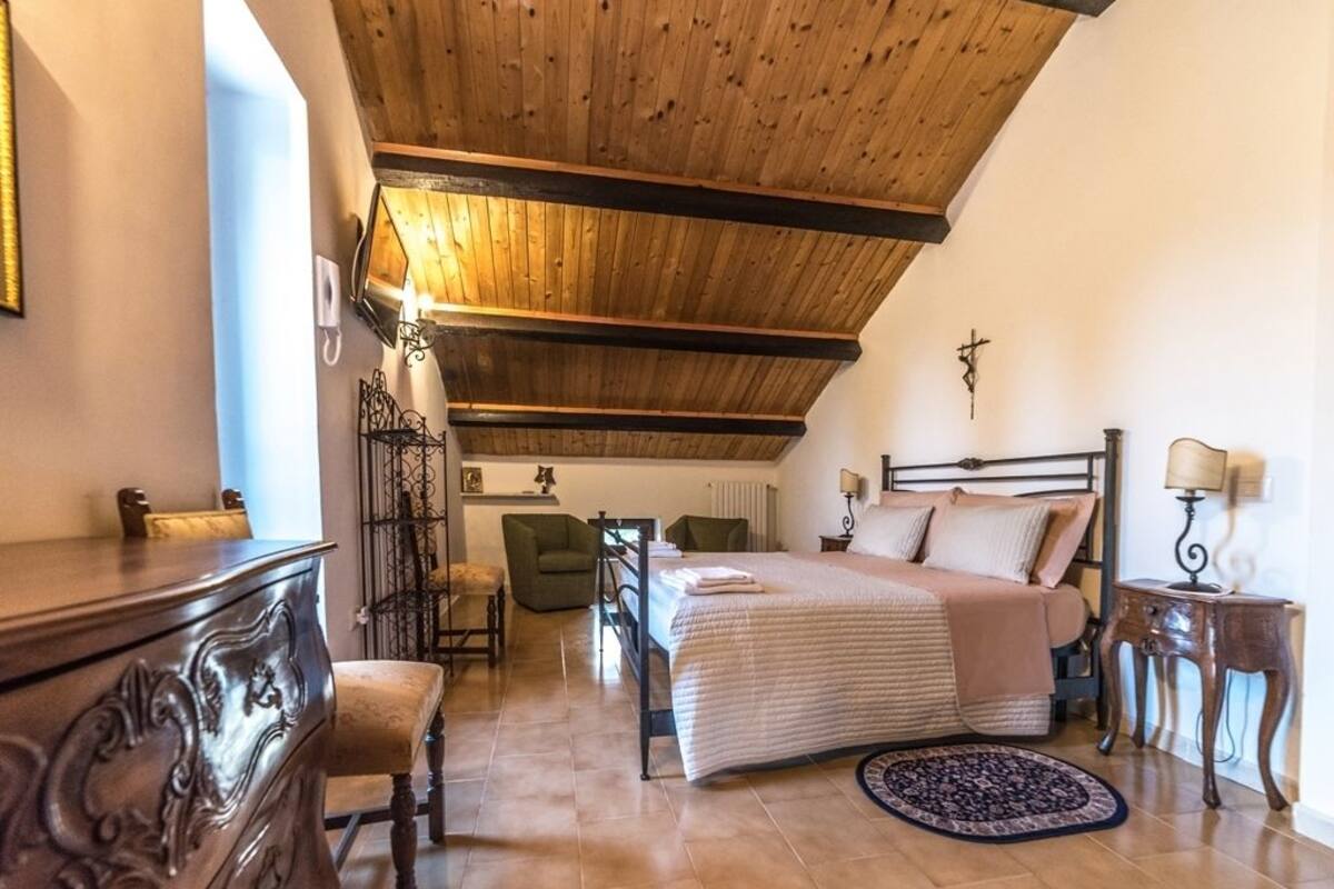 Um quarto com teto de madeira, uma cama com cabeceira, mesa de cabeceira, uma cadeira e um aparador.