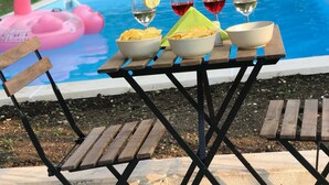 Mesa à beira da piscina com tigelas de batata e copos de vinho, cercada por um deck de madeira e um flutuador de flamingo rosa.