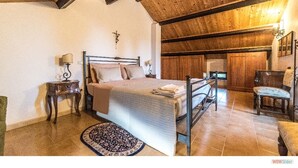 Quarto com teto de madeira, uma cama com colcha branca, mesa de cabeceira com luminária e uma cadeira.