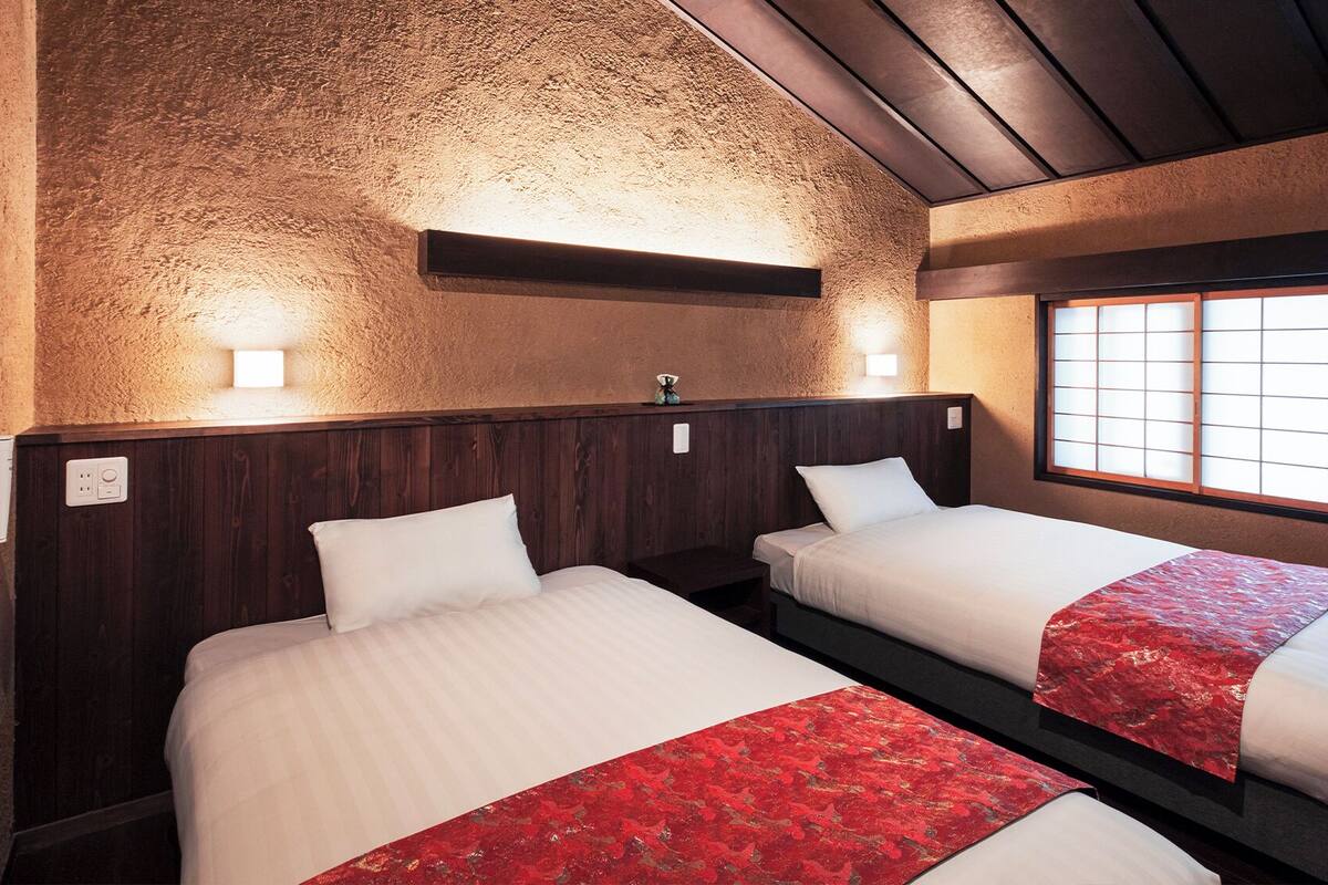 Quarto de hotel com duas camas, parede texturizada, teto de madeira e janela com padrão quadriculado.