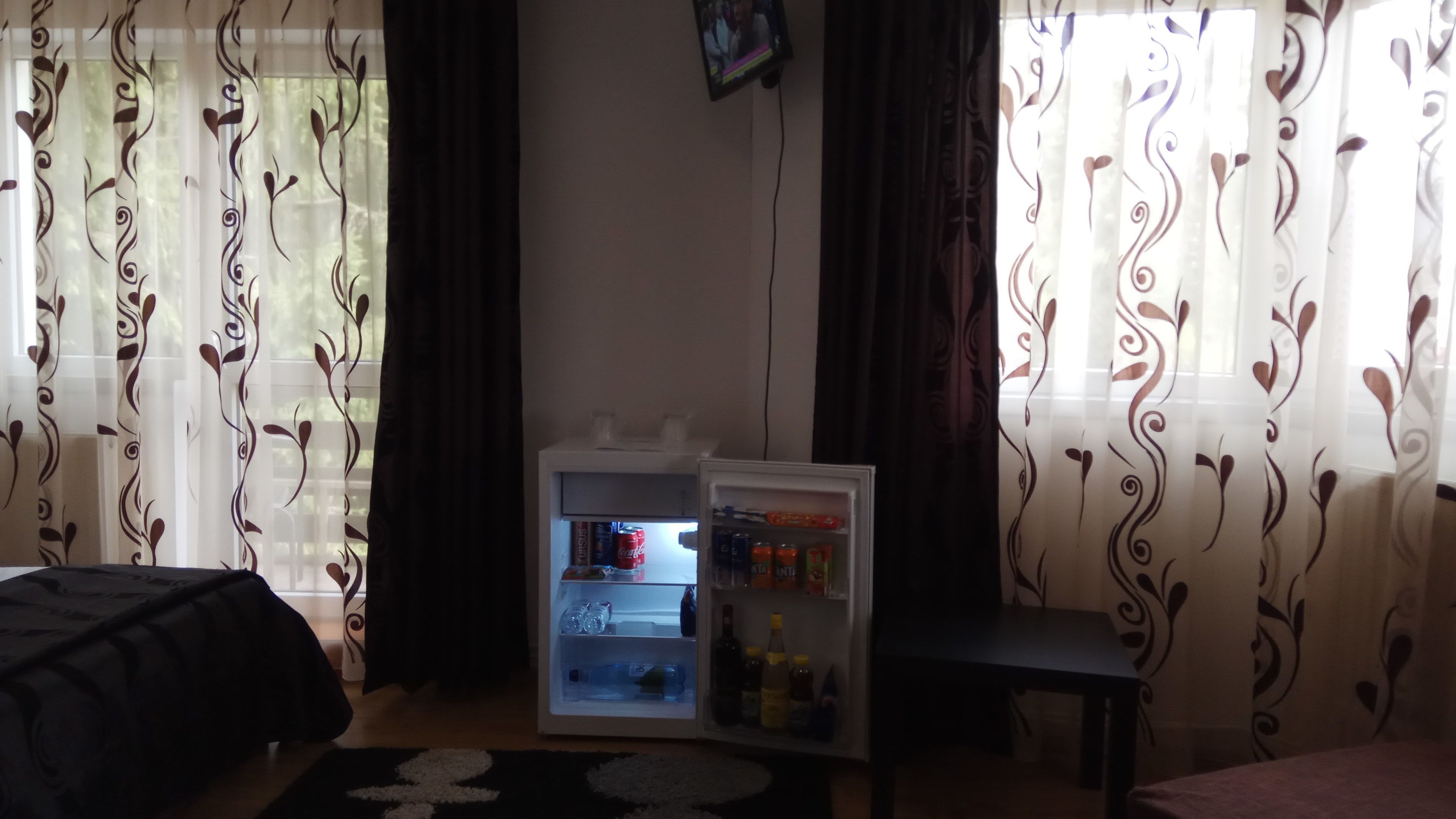 Quarto pequeno com geladeira repleta de bebidas e acompanhamentos, cama com cobertura escura e cortinas estampadas com folhas.