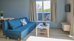 Uma sala de estar moderna com um sofá azul, uma pequena mesa de madeira, uma TV fixada na parede e uma janela com cortinas.