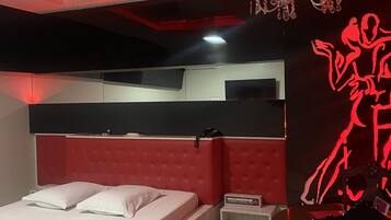 Um quarto moderno com esquema de cores vermelho e preto, uma cama com roupa de cama branca, uma mesa de cabeceira com abajur e uma TV fixada na parede.
