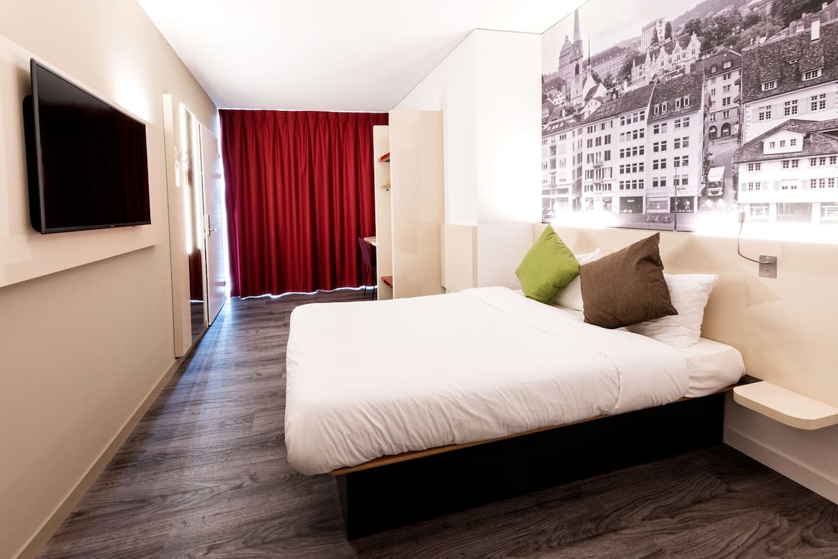 Quarto de hotel moderno com uma cama grande, televisão de tela plana na parede, cortina vermelha e vista para a cidade.