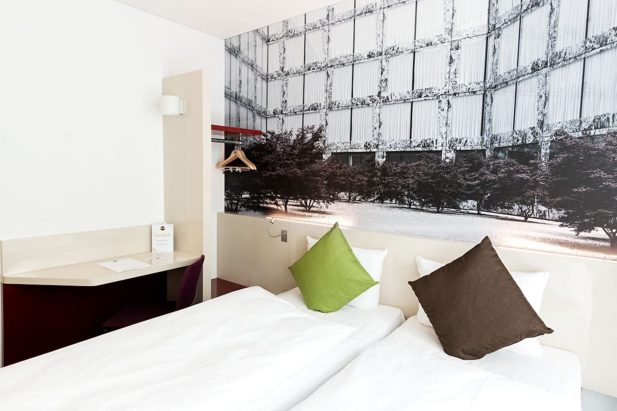Quarto de hotel com uma cama, uma escrivaninha e uma janela grande com vista para árvores e um edifício.