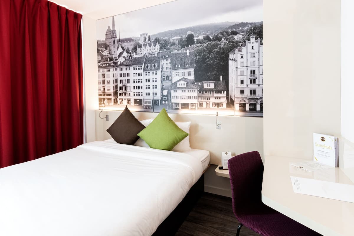 Quarto de hotel com cama, travesseiros, uma cadeira, janela e vista panorâmica da cidade, além de cortina vermelha.