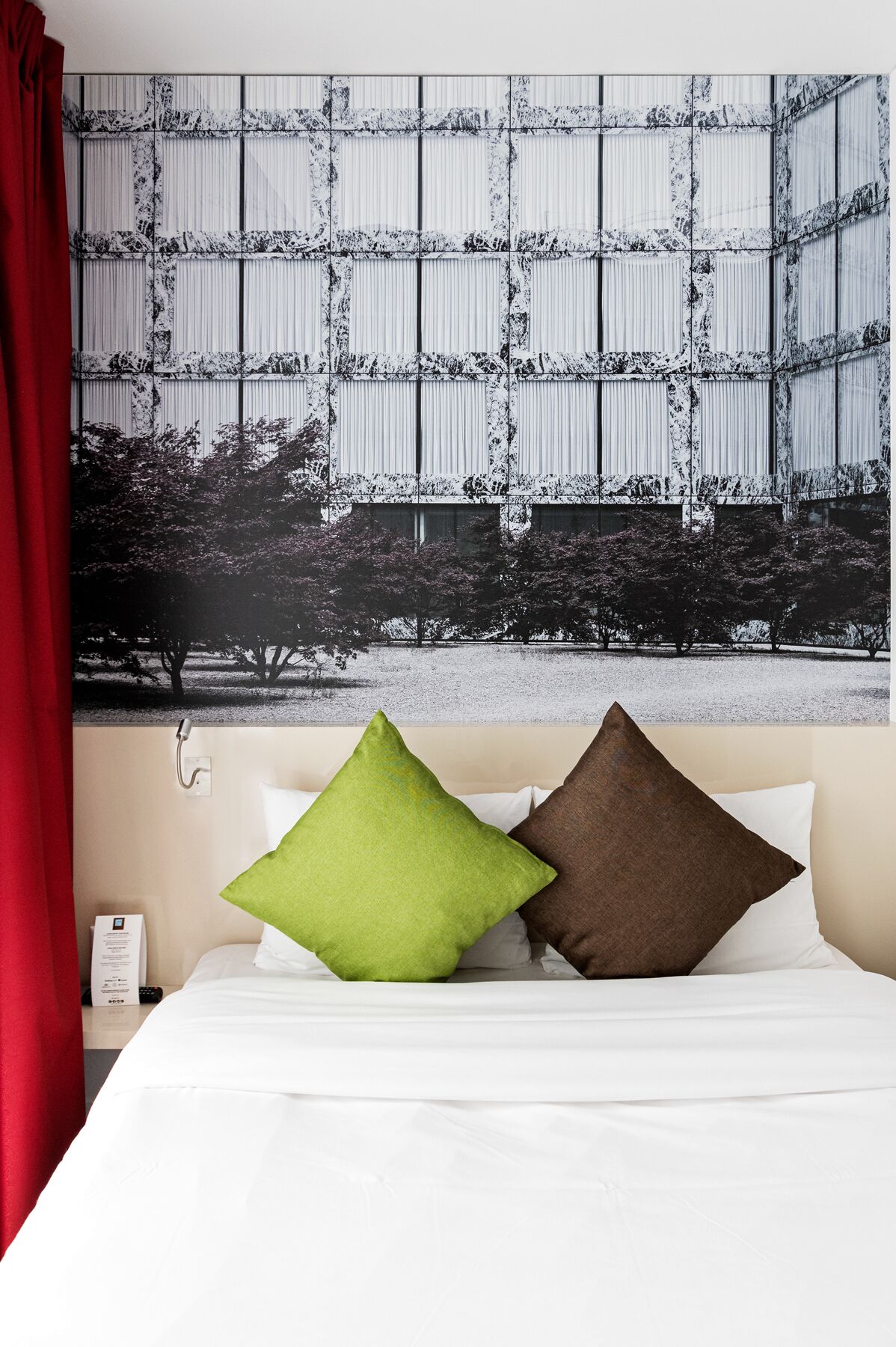 Quarto de hotel com uma cama, duas almofadas, uma mesinha de cabeceira, uma cortina vermelha e uma grande obra de arte na parede retratando árvores e um edifício.