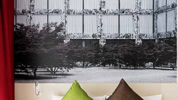 Quarto de hotel com uma cama, duas almofadas, uma mesinha de cabeceira, uma cortina vermelha e uma grande obra de arte na parede retratando árvores e um edifício.