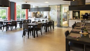 Uma área de jantar moderna com várias mesas e cadeiras, uma cozinha ao fundo e janelas amplas com cortinas vermelhas.