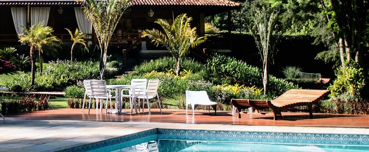 Uma área junto à piscina com mesa e cadeiras, cercada por vegetação exuberante e um deck de madeira.