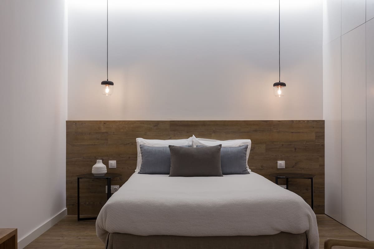 Quarto moderno com cabeceira de madeira, uma cama grande com roupa de cama branca, duas mesas de cabeceira e luminárias pendentes.