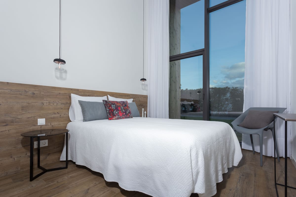 Quarto moderno com uma cama grande, uma mesinha, uma cadeira e uma janela com cortinas.