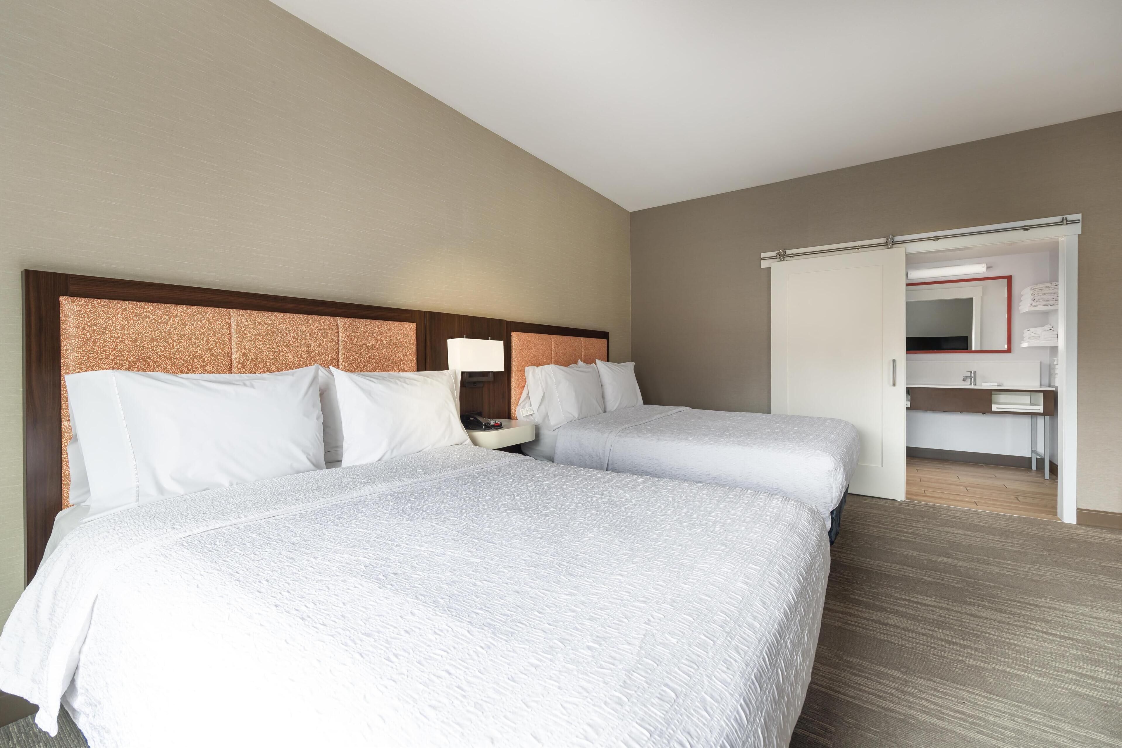 Quarto de hotel com duas camas, uma cabeceira de madeira, mesa de cabeceira com abajur e armário com espelho e prateleiras.