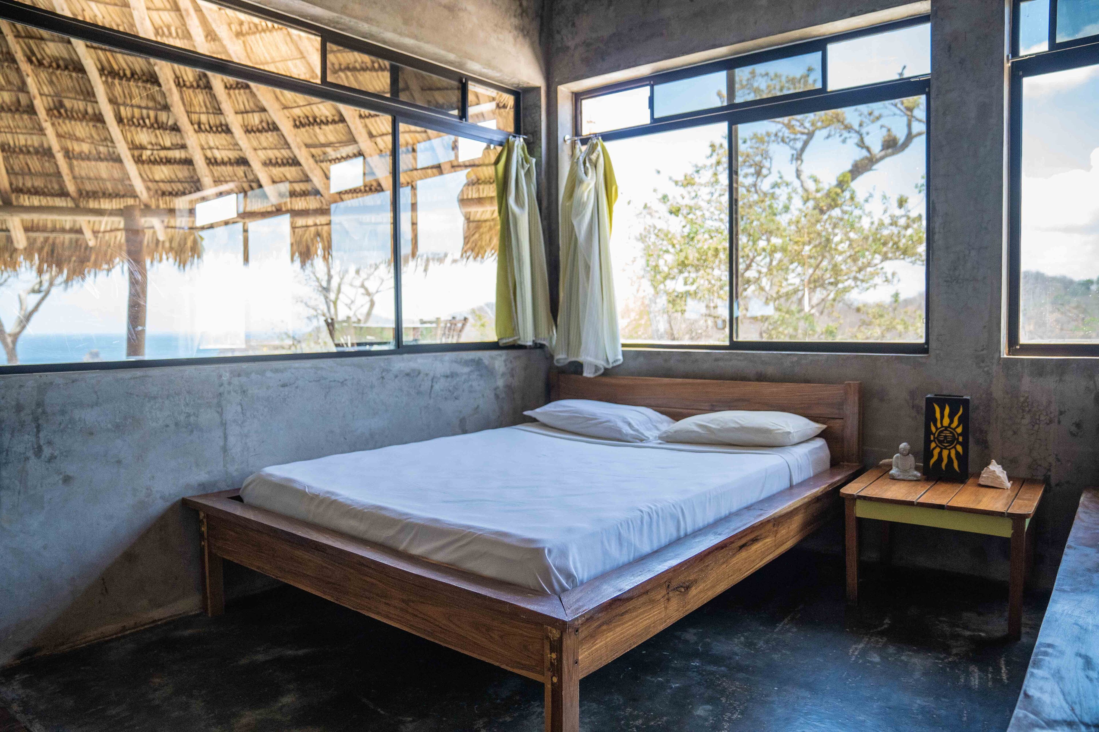 Quarto com cama de madeira, pequena mesa de cabeceira e janelas amplas com vista para árvores e céu.