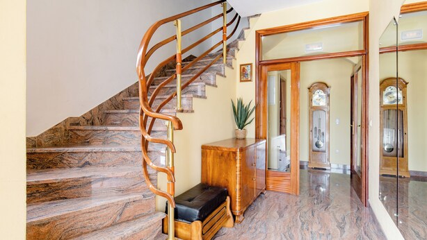 Uma escadaria de mármore com guarda-mão de madeira, um banco de madeira, um armário e uma porta que dá para um quarto com um relógio e um espelho.