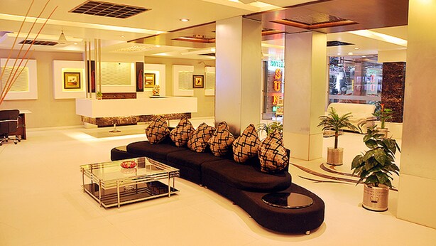 Um lobby moderno com um sofá preto decorado com almofadas estampadas, uma mesa de centro de vidro e plantas em vasos.