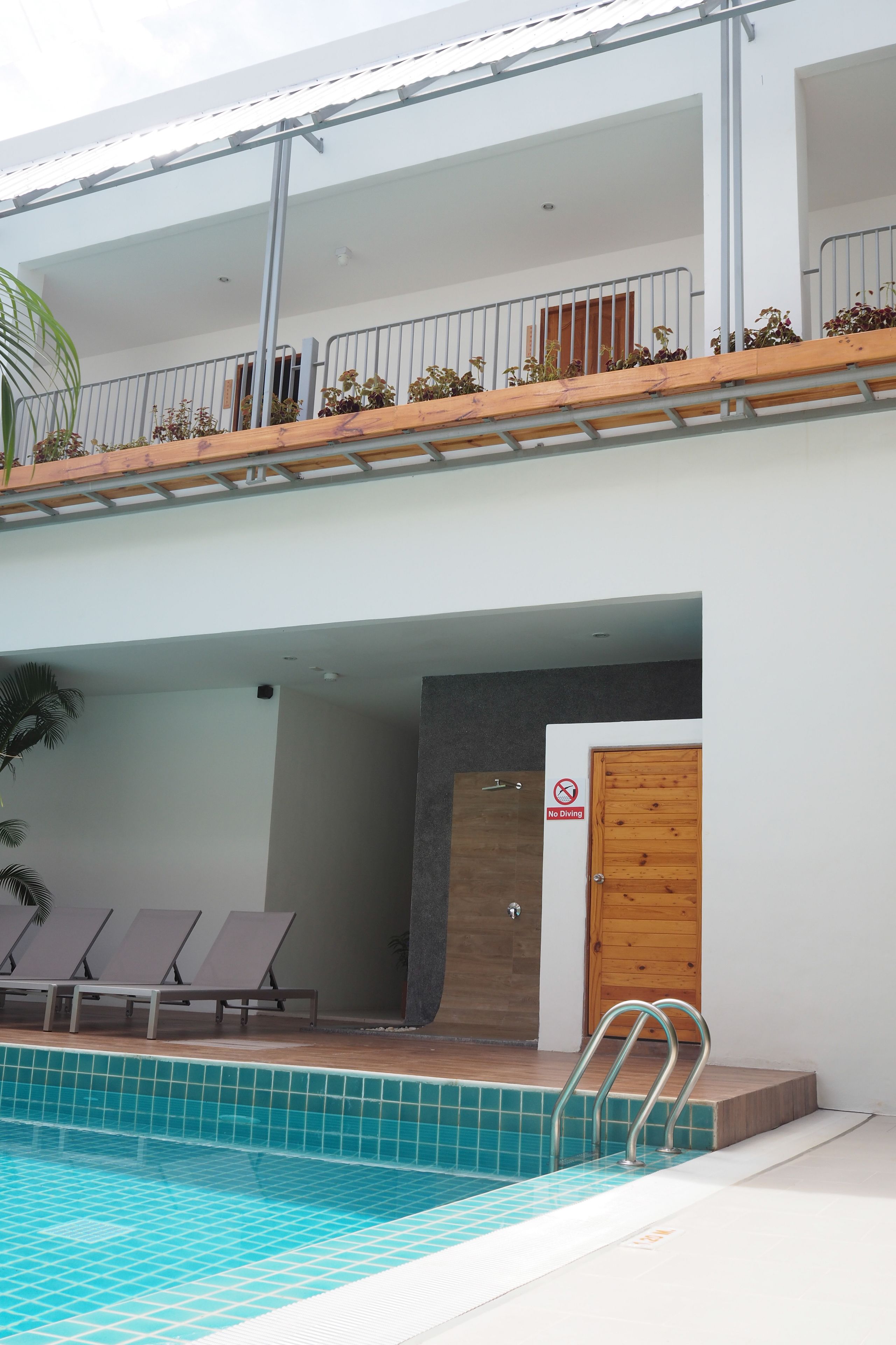 Piscina com borda azulejada, porta de madeira e varanda com grades de metal e plantas.
