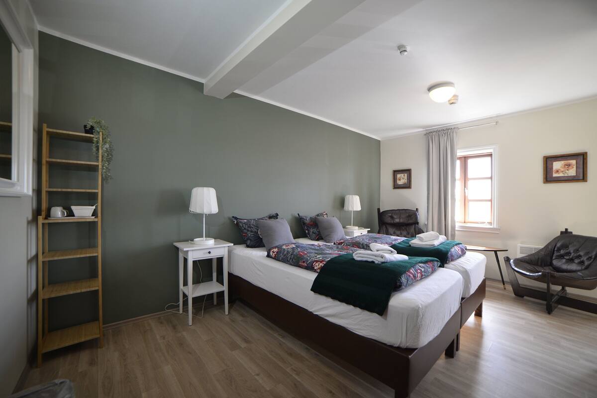 Quarto com duas camas, uma estante de madeira, uma escrivaninha, uma cadeira e uma janela com cortinas.