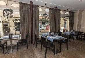 Uma área de refeições com mesas preparadas para dois, grandes janelas com cortinas e vista para a rua.