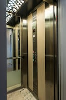 Interior de elevador com paredes metálicas, painel de madeira e painel de controle com botões e visor digital.