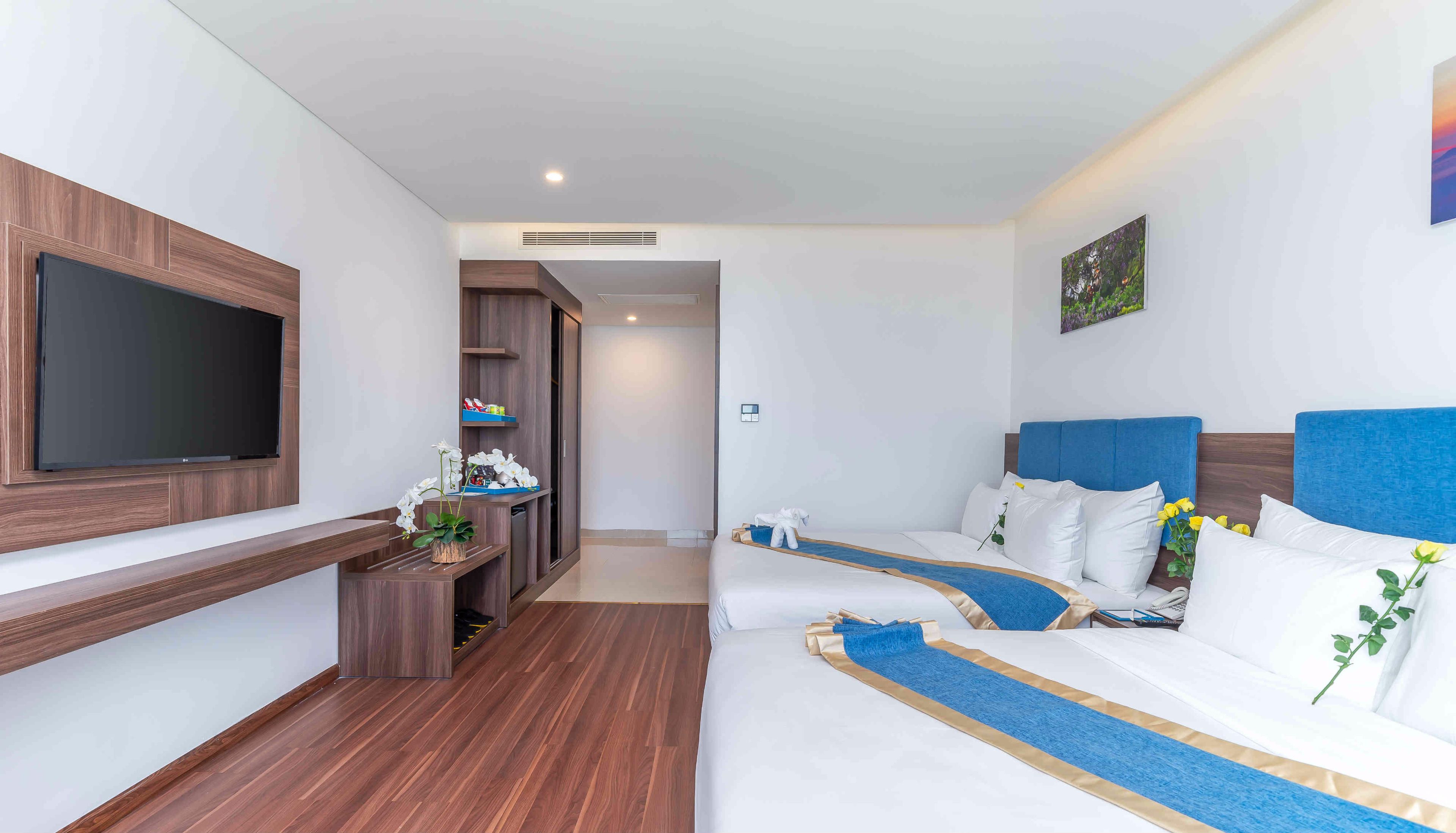 Quarto de hotel com televisão de tela grande, cama com cabeceira azul, roupa de cama branca e uma pequena mesa de cabeceira.