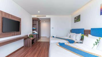 Quarto de hotel com televisão de tela grande, cama com cabeceira azul, roupa de cama branca e uma pequena mesa de cabeceira.