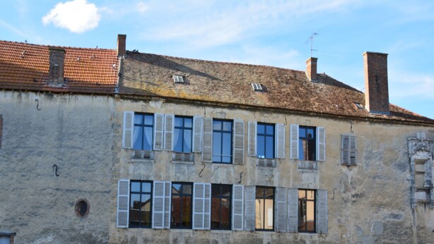 Um edifício histórico com várias janelas e venezianas, fachada de pedra e um rio ao primeiro plano.