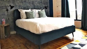 Quarto moderno com cama azul, roupa de cama branca e almofadas estampadas. Há uma pequena mesa de cabeceira de madeira com abajur e um objeto decorativo. O quarto tem uma parede de concreto e uma janela grande com cortinas.