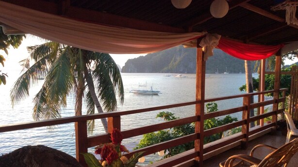 Um deck de madeira com vista para o mar, palmeiras e um barco ao longe.