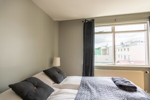Cama bem arrumada com edredom branco e cinza, duas almofadas pretas e uma toalha dobrada no lado direito. Um abajur branco está do lado esquerdo da cama, e uma janela mostra vista de prédios e um telhado vermelho.