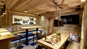 Um interior acolhedor e rústico, com teto de madeira, detalhes em pedra, uma televisão grande fixada na parede e um espaço bar com banquetas e mesa de madeira.
