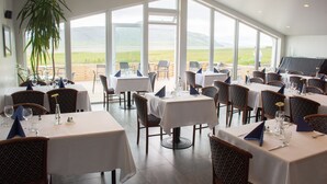 Restaurante com mesas preparadas para refeições, grandes janelas com vista para o exterior e uma planta no canto.