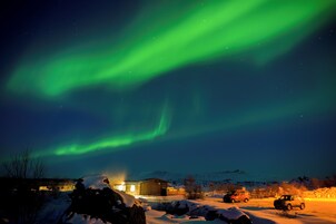 Paisagem nevada com um edifício e carros estacionados sob um céu noturno com a aurora boreal.