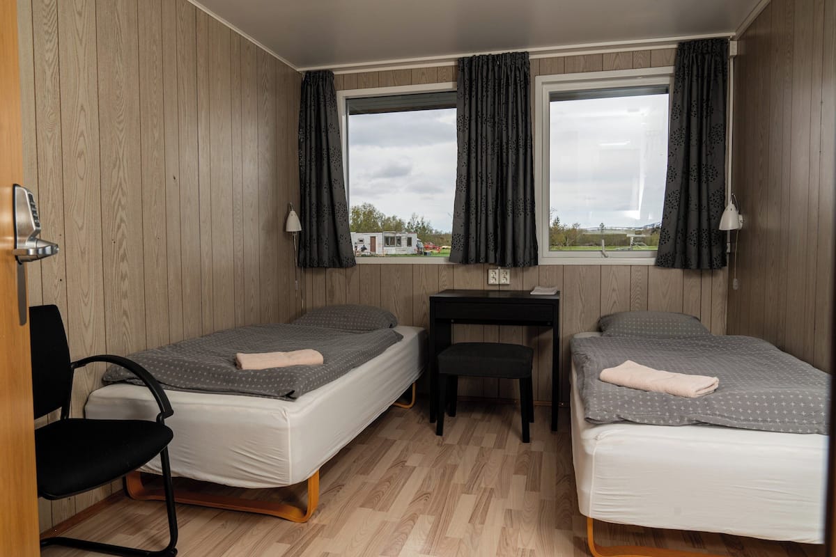Quarto com duas camas de solteiro, uma escrivaninha, uma cadeira e janela com cortinas.