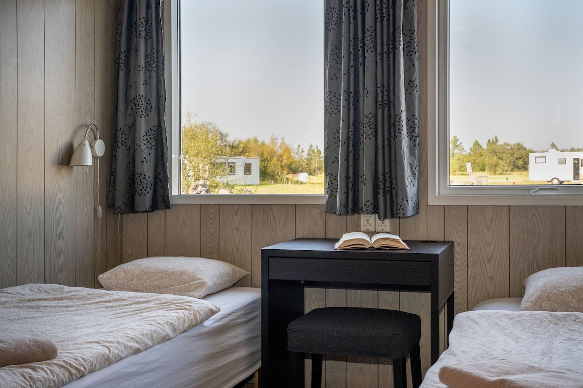 Quarto com duas camas, uma mesinha com um livro aberto e vista para uma caravana e árvores lá fora.
