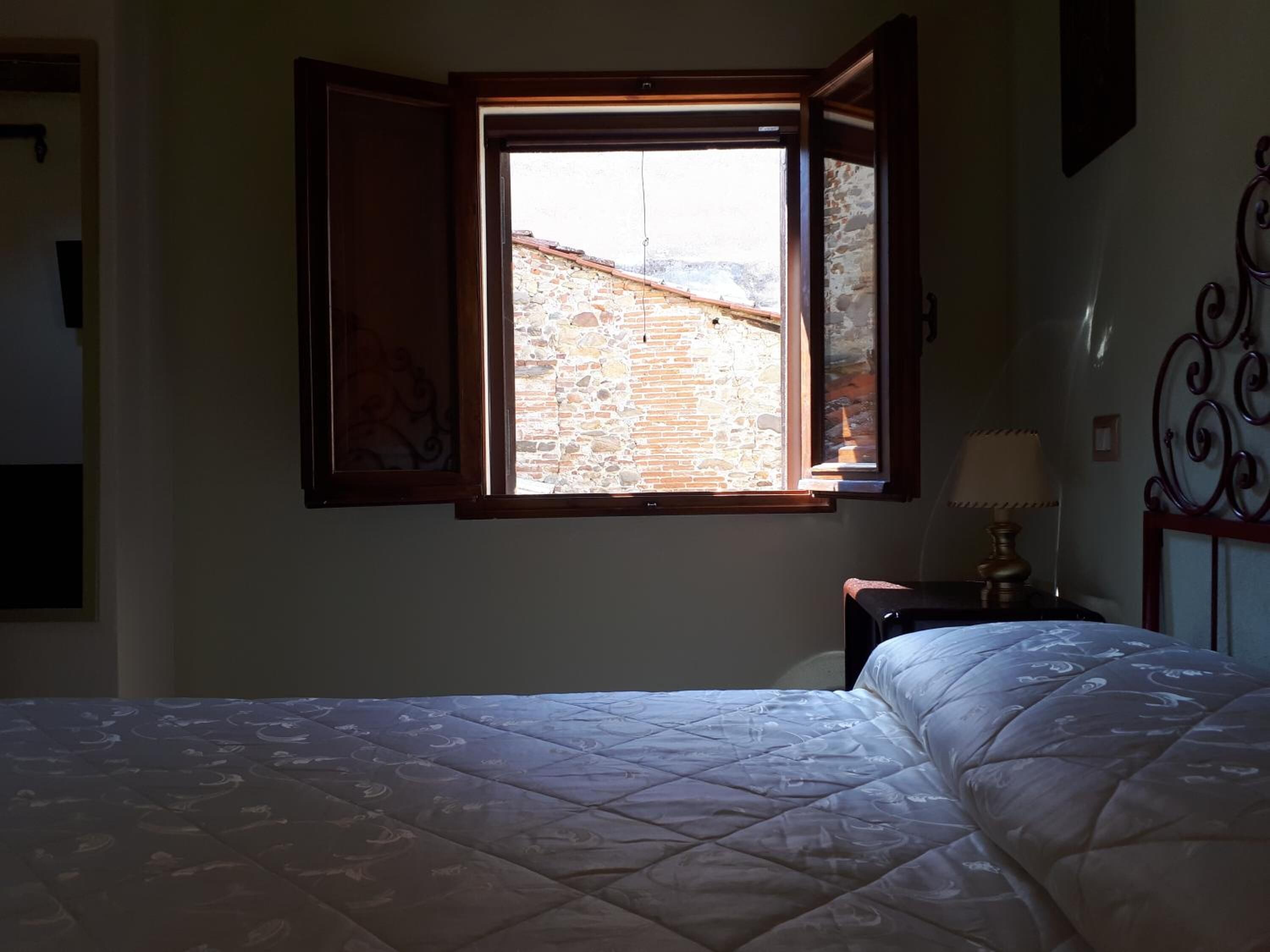 Quarto com cama, mesa de cabeceira, luminária e janela com persiana aberta.