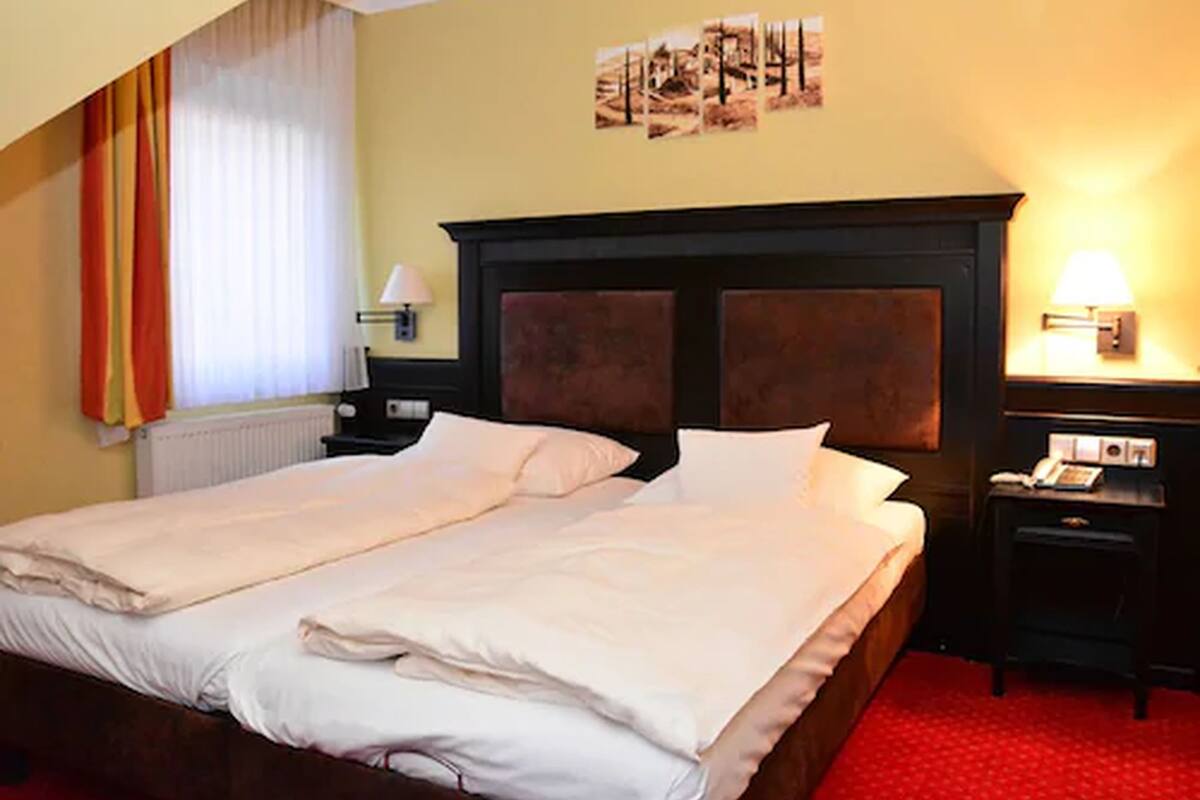 Uma cama de casal com lençóis brancos, cabeceira de madeira escura, mesa de cabeceira com abajur e telefone fixado na parede.