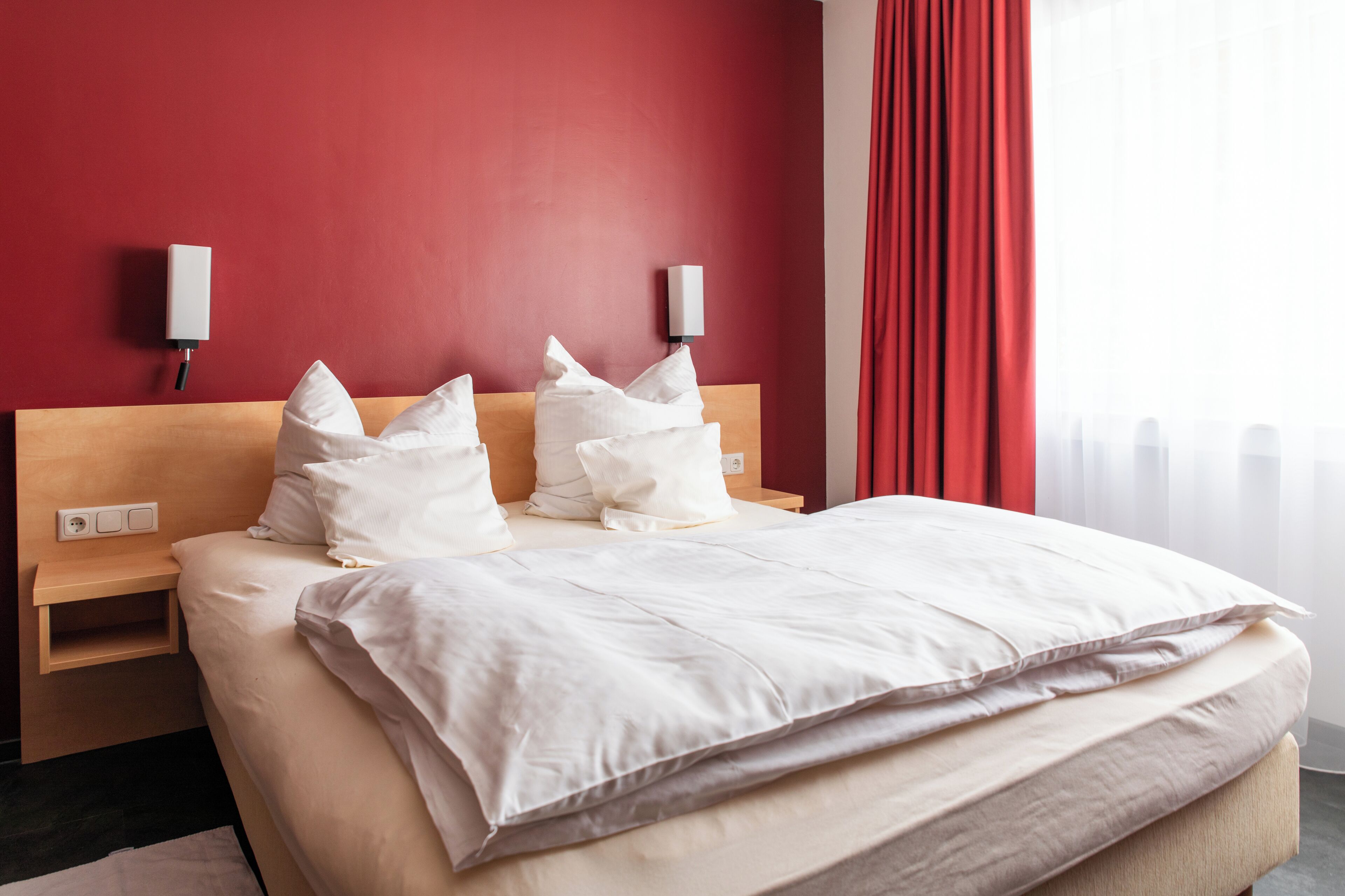 Cama bem arrumada com lençóis e almofadas brancas, cabeceira de madeira, mesas de cabeceira com abajures e parede vermelha com janela e cortinas.
