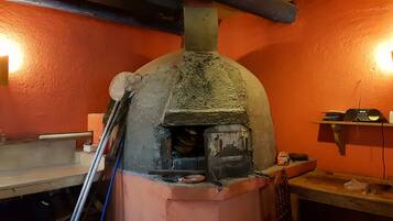 Forno de tijolos com chaminé, um esfregão encostado e uma prateleira com rádio e livros.