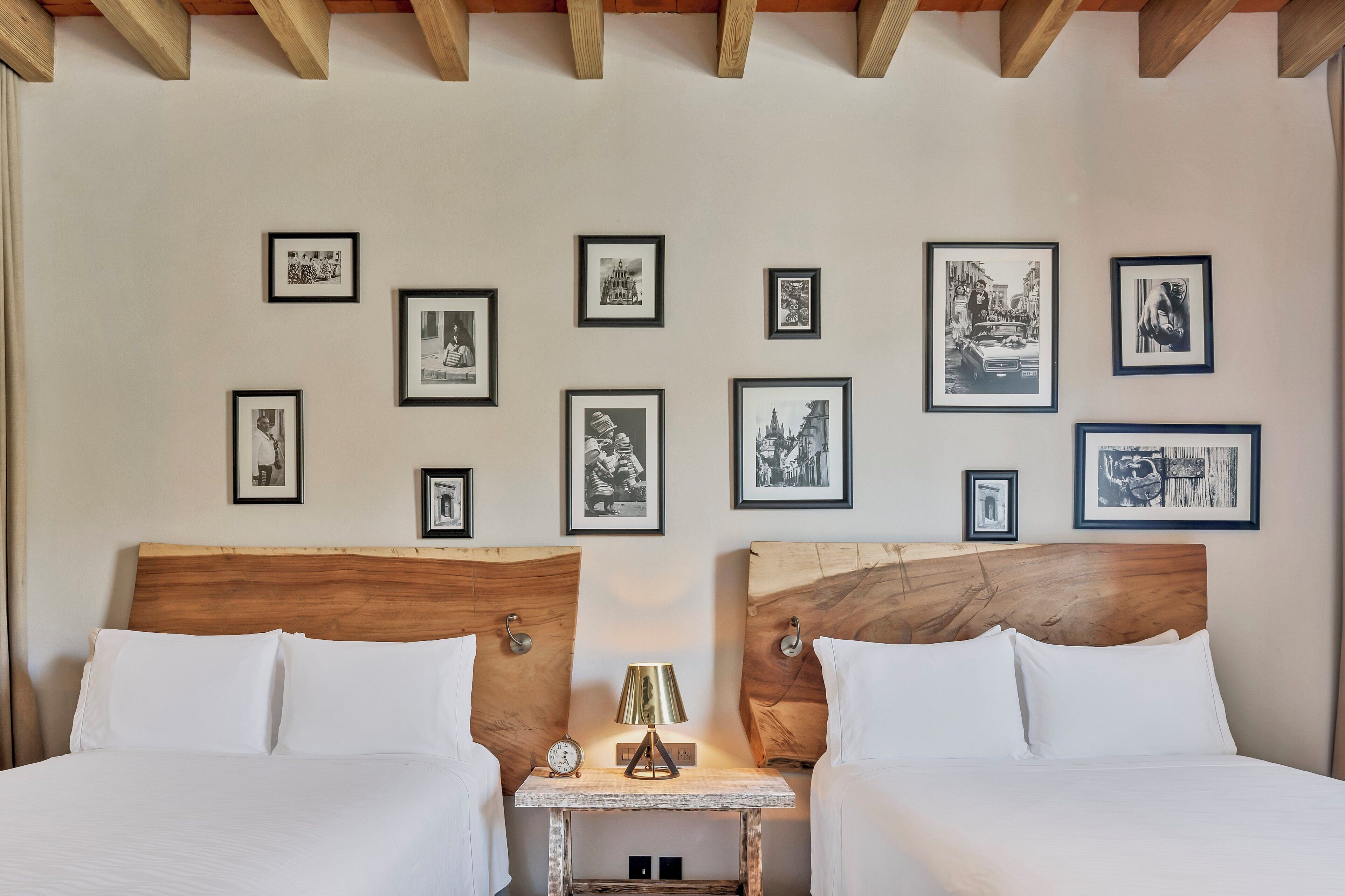 Quarto de hotel com duas camas, uma mesinha com abajur e parede decorada com fotografias em preto e branco.