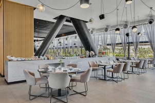 Interior moderno de restaurante com mesas redondas, cadeiras e um balcão central com comida e bebidas.