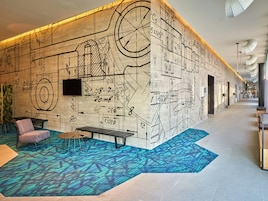 Um saguão de hotel com uma grande parede de desenhos arquitetônicos, área de estar com carpete estampado e um corredor longo com design moderno.