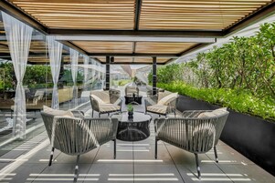 Área externa com mobiliário de vime, uma mesa redonda central e um espaço envidraçado com vista para o verde ao redor.