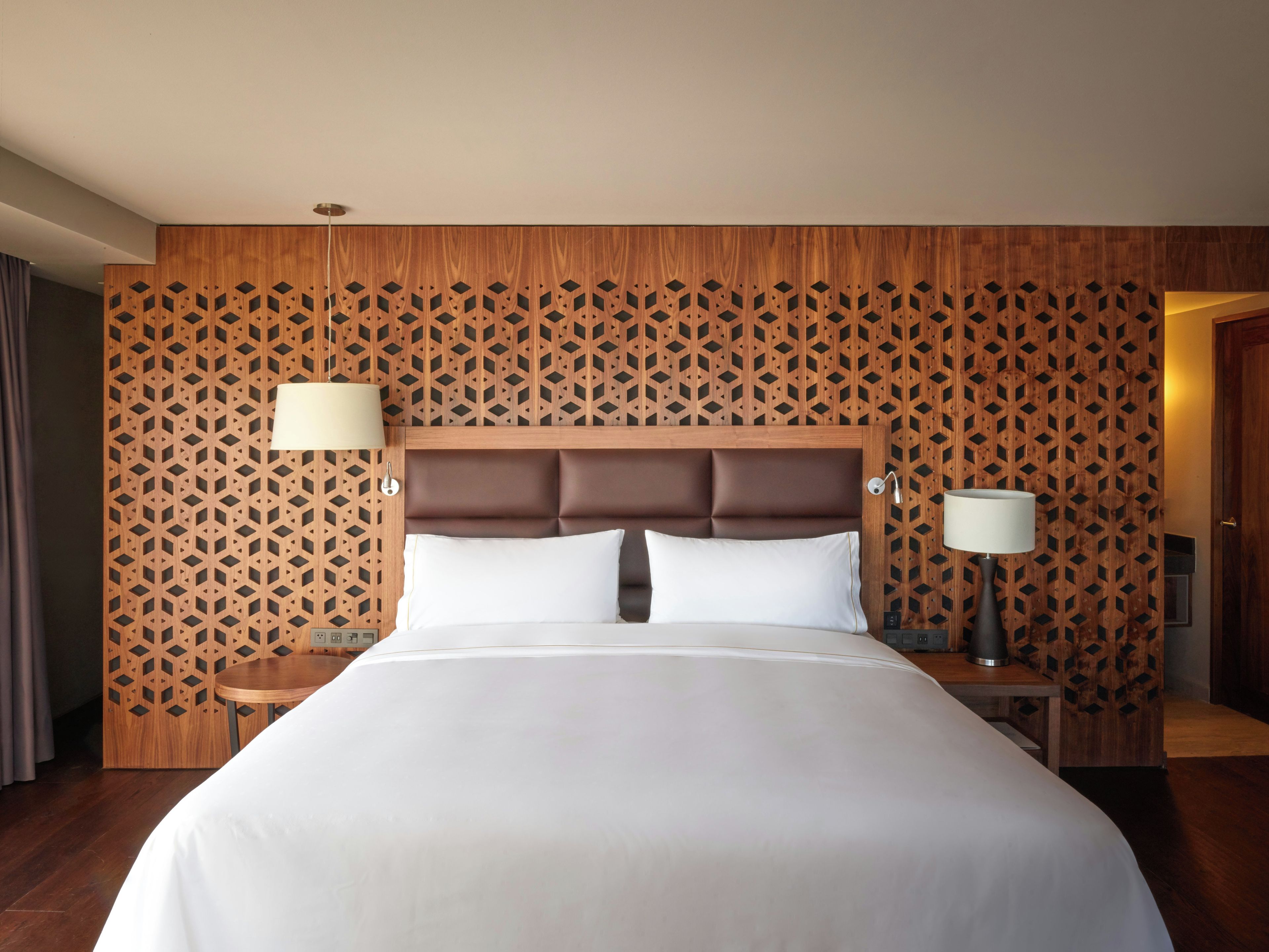 Quarto de hotel com uma cama grande, cabeceira de madeira, duas luminárias laterais e parede estampada.
