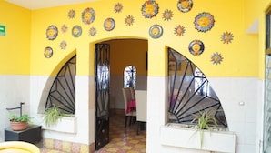 Quarto com paredes amarelas, chão de azulejos, janela com moldura preta e porta com arco de metal decorativo. Há plantas em vasos no chão e uma mesa com uma cadeira.