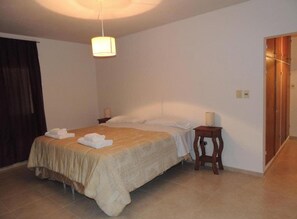 Quarto de hotel com uma cama, mesa de cabeceira com abajur, guarda-roupa de madeira e uma porta.