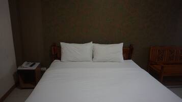 Quarto de hotel com uma cama grande, uma mesinha de cabeceira de madeira com telefone e uma cadeira de madeira.