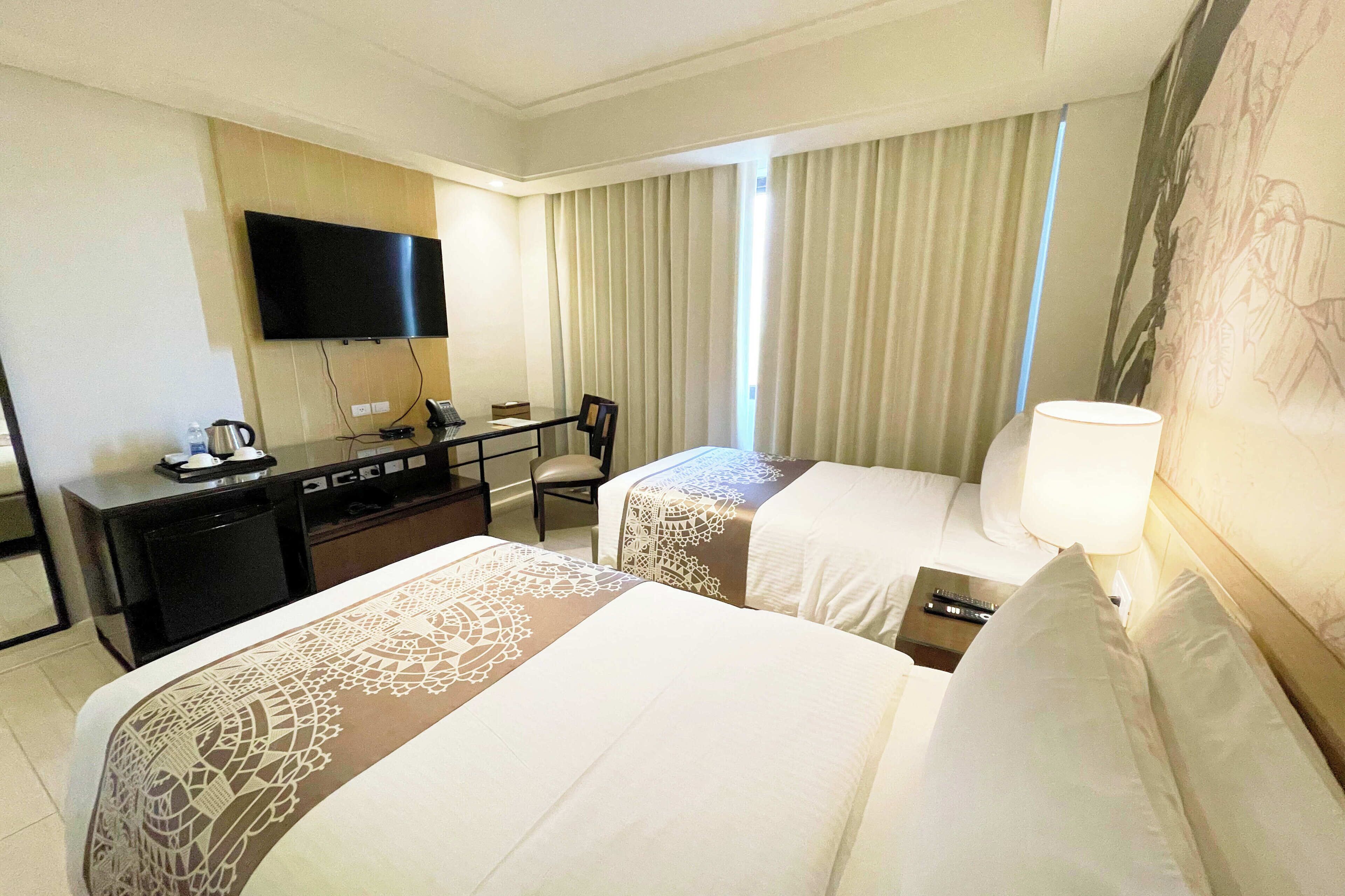 Quarto de hotel com duas camas, uma escrivaninha, televisão e janela com cortinas.