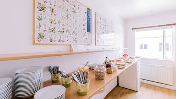 Espaço de buffet de café da manhã com diversas opções de alimentos e utensílios, um quadro de avisos com ilustrações de plantas e uma janela que proporciona luz natural.