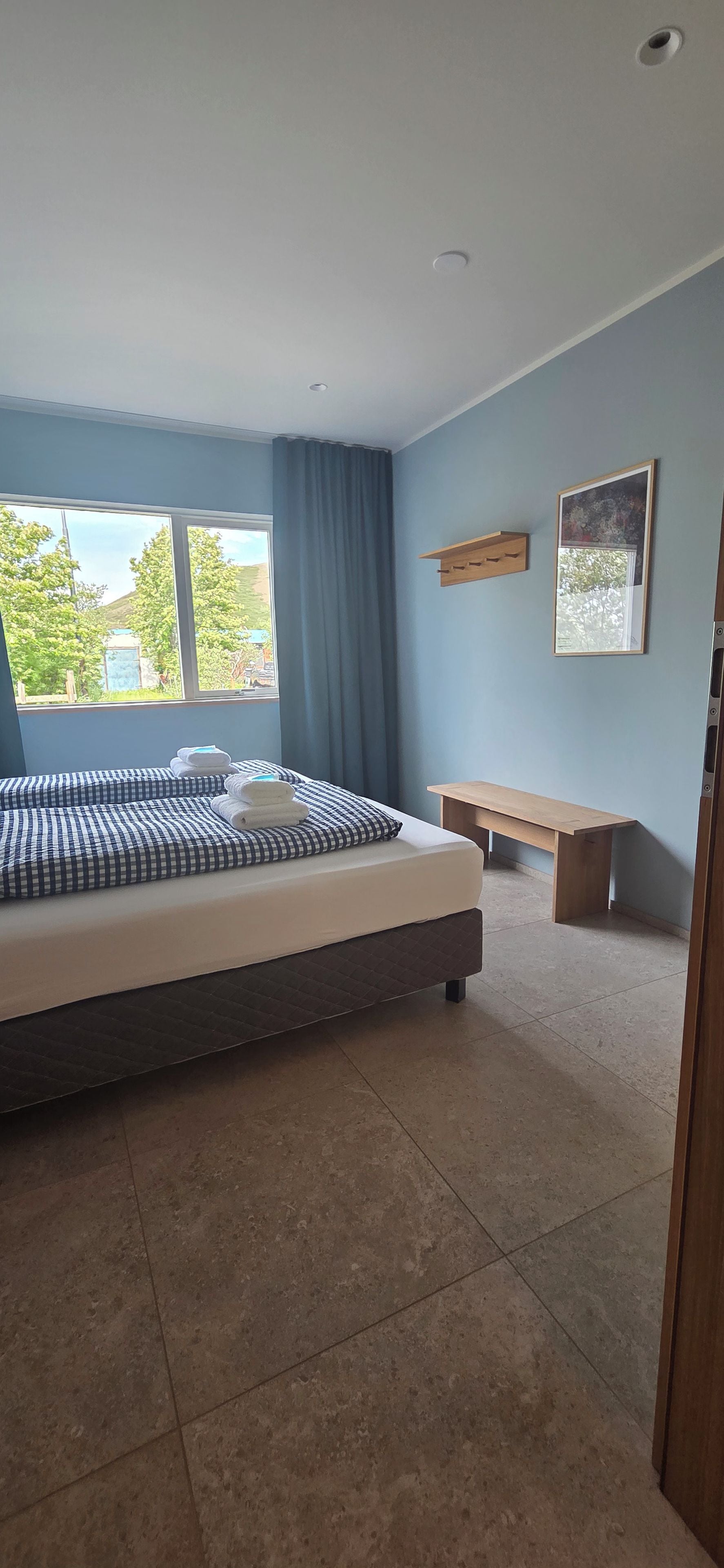 Quarto com cama, um banquinho, janela com vista e prateleira na parede.